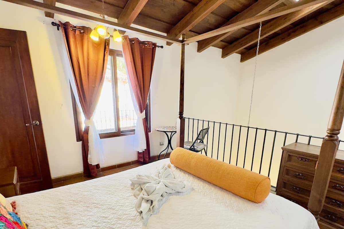 Quarto com uma cama grande, vigas de madeira no teto, uma mesa pequena e uma cadeira.