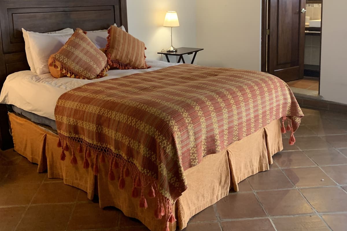 Quarto com cama grande, cabeceira de madeira, mesa de cabeceira com abajur e porta para o banheiro.