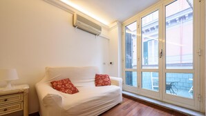 Quarto com sofá branco, mesa de cabeceira com abajur e grande janela com vista para o exterior.