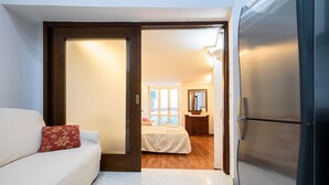 Um espaço compacto com sofá branco, almofada vermelha com flores, cama com colcha branca, piso de madeira e uma moderna geladeira de aço inoxidável.