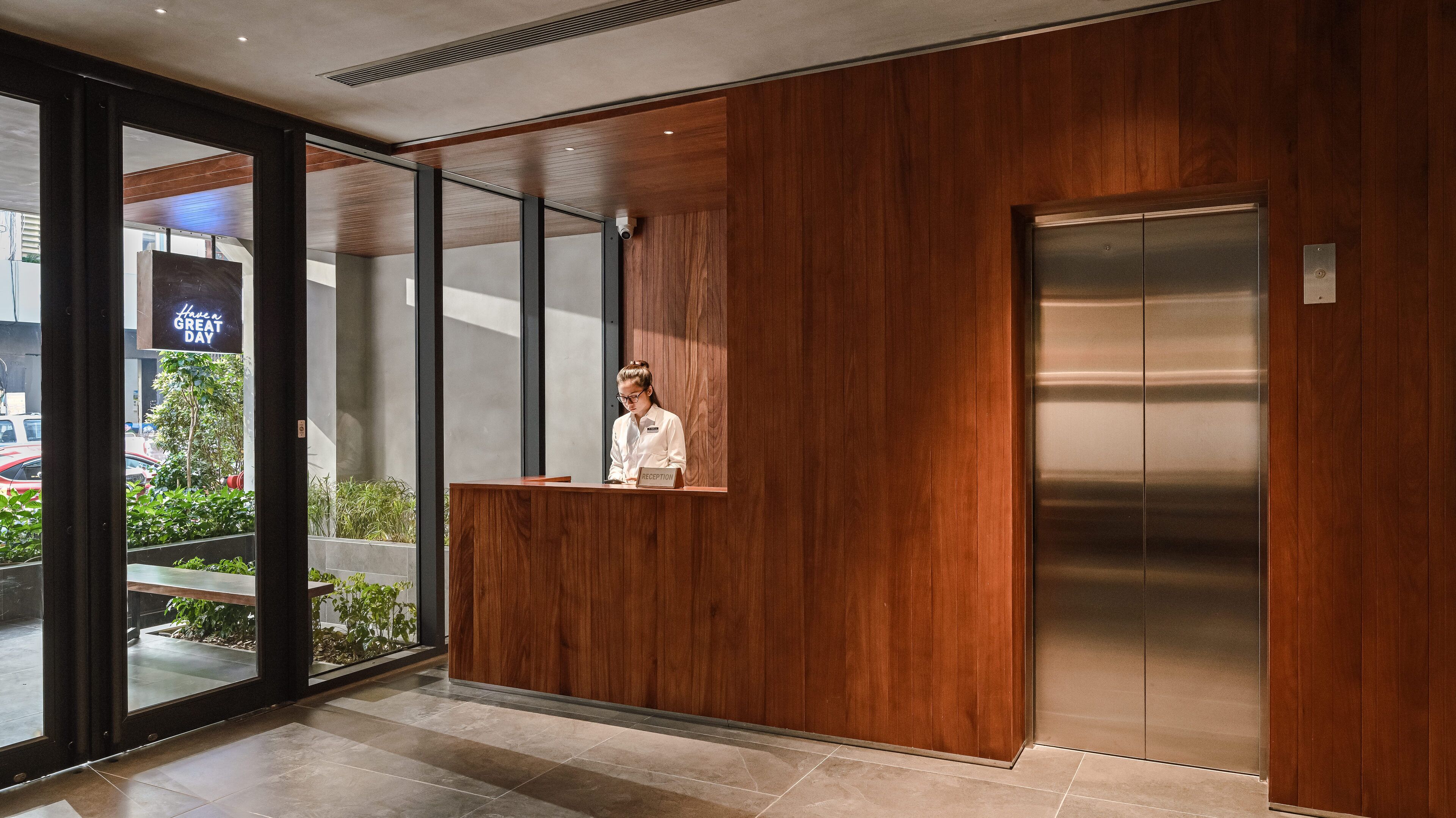 Bancada de recepção com acabamento em madeira, porta de vidro para o exterior e um elevador moderno.