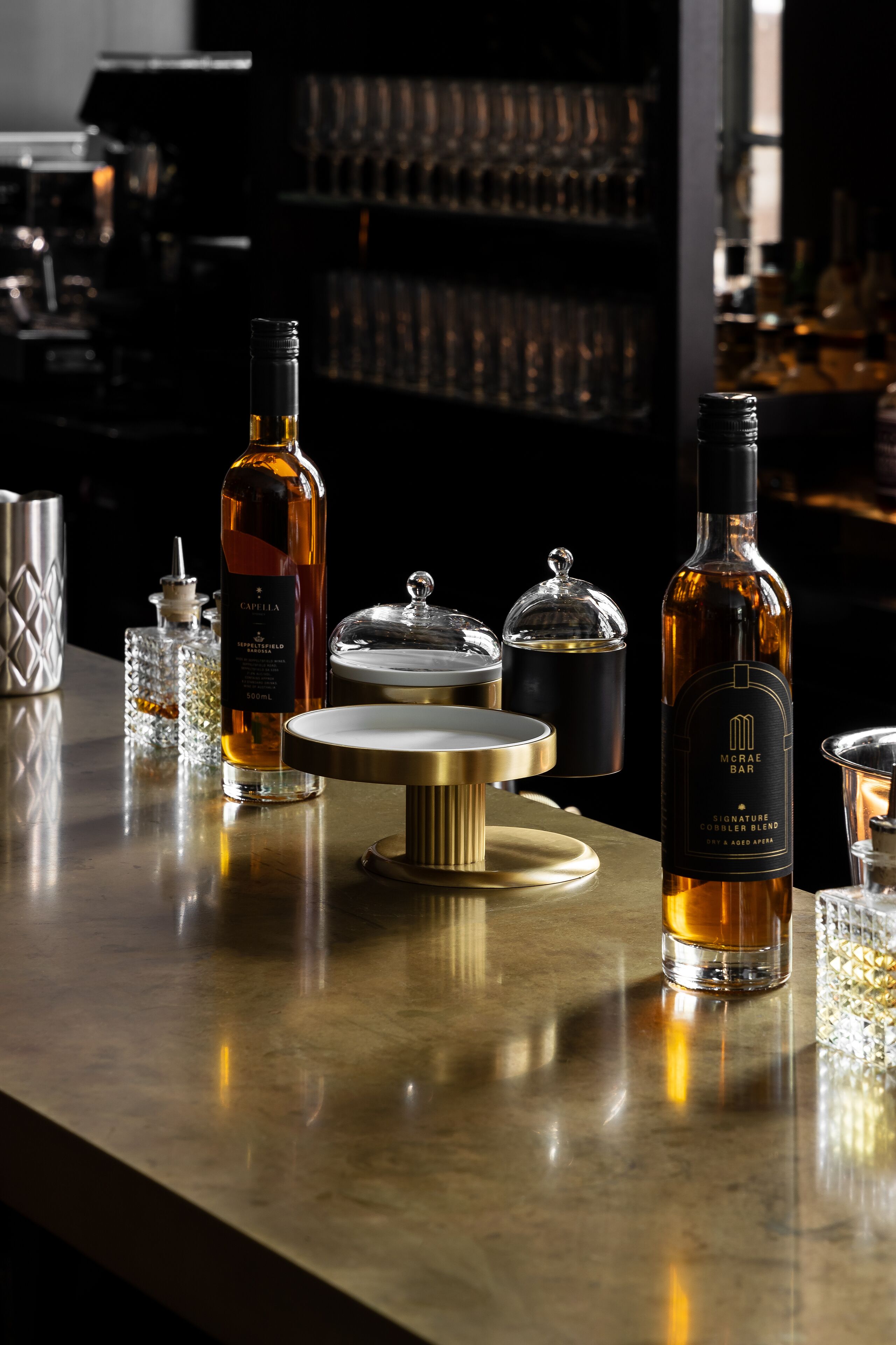 Uma área de bar com garrafas de bebidas, um copo e um recipiente de metal sobre uma superfície refletora.