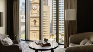 Quarto de hotel com uma janela ampla que oferece vista para um campanário e edifícios altos. Há uma mesa redonda com livros e uma luminária.