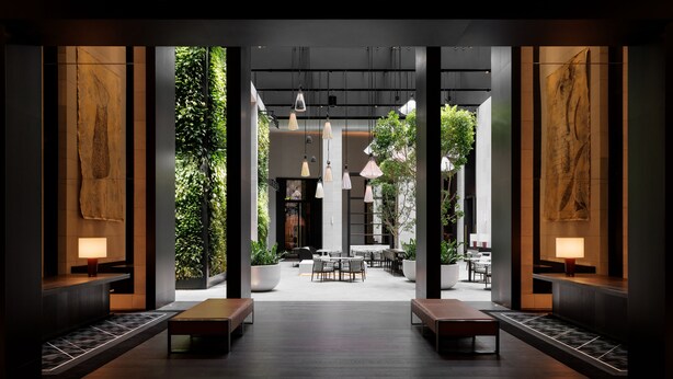 Interior moderno com espaço aberto central, bancos, luminárias pendentes e vista para área externa com plantas e mesas.