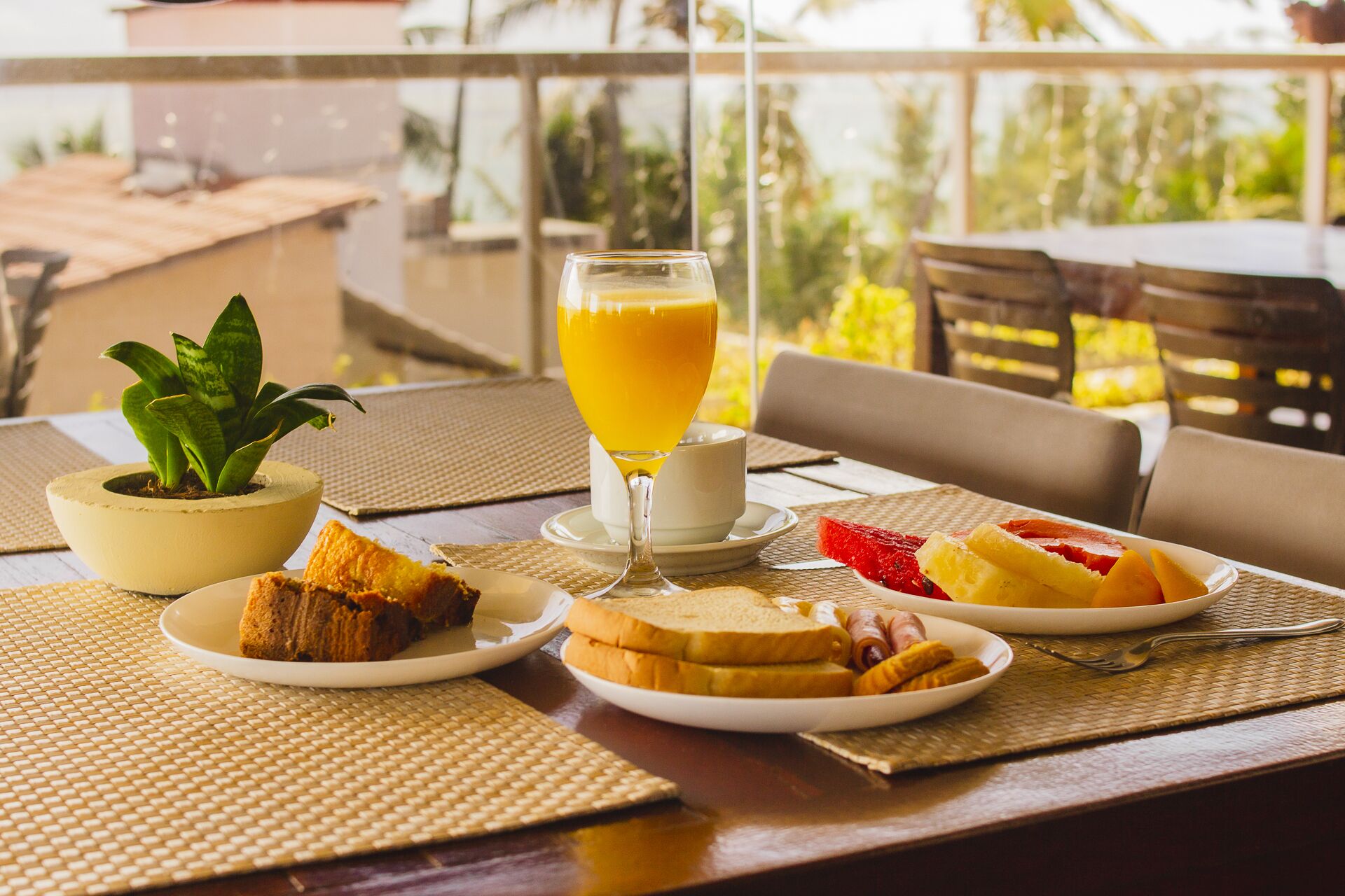 Disposição para o café da manhã com uma planta, copo de suco de laranja, torradas e frutas sobre uma mesa.