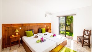 Quarto com duas camas, cabeceira de madeira, mesas de cabeceira com abajures e vista para um jardim através de uma porta deslizante.