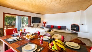 Uma sala de jantar com mesa de madeira posta para refeição, um sofá com almofadas, uma lareira e uma televisão.