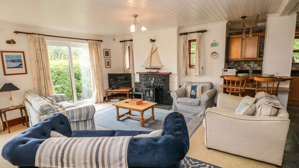 Uma sala de estar acolhedora com um sofá azul, uma poltrona listrada e um sofá bege. Há uma lareira, uma televisão e um espaço de jantar com cadeiras de madeira.