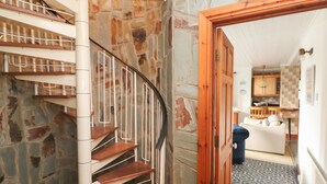 Uma escadaria em espiral com paredes de pedra, uma porta de madeira que dá para uma sala de estar com cozinha, e um tapete estampado.