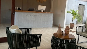 Uma área de recepção com balcão moderno, luminárias pendentes de vime e um espaço de estar com cadeiras pretas e mesa redonda.