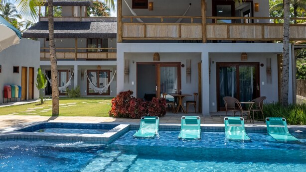Resort de dois andares com piscina, palmeiras e estrutura com telhado de palha.
