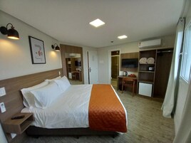 Quarto de hotel com cama, mesa de cabeceira, luminária, guarda-roupa e escrivaninha com cadeira.