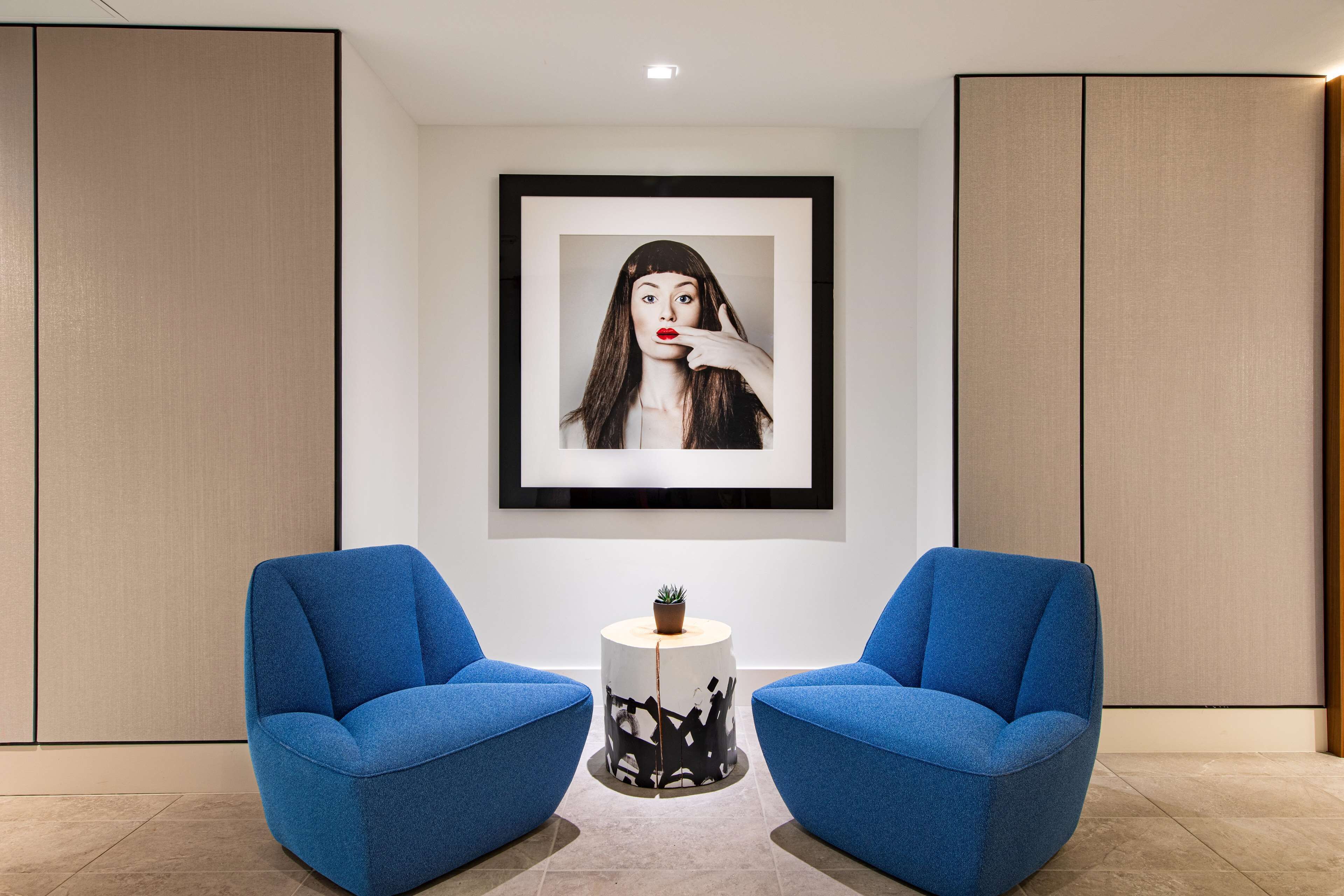 Quarto moderno com duas poltronas azuis, uma mesinha redonda com uma planta e uma obra de arte emoldurada na parede.