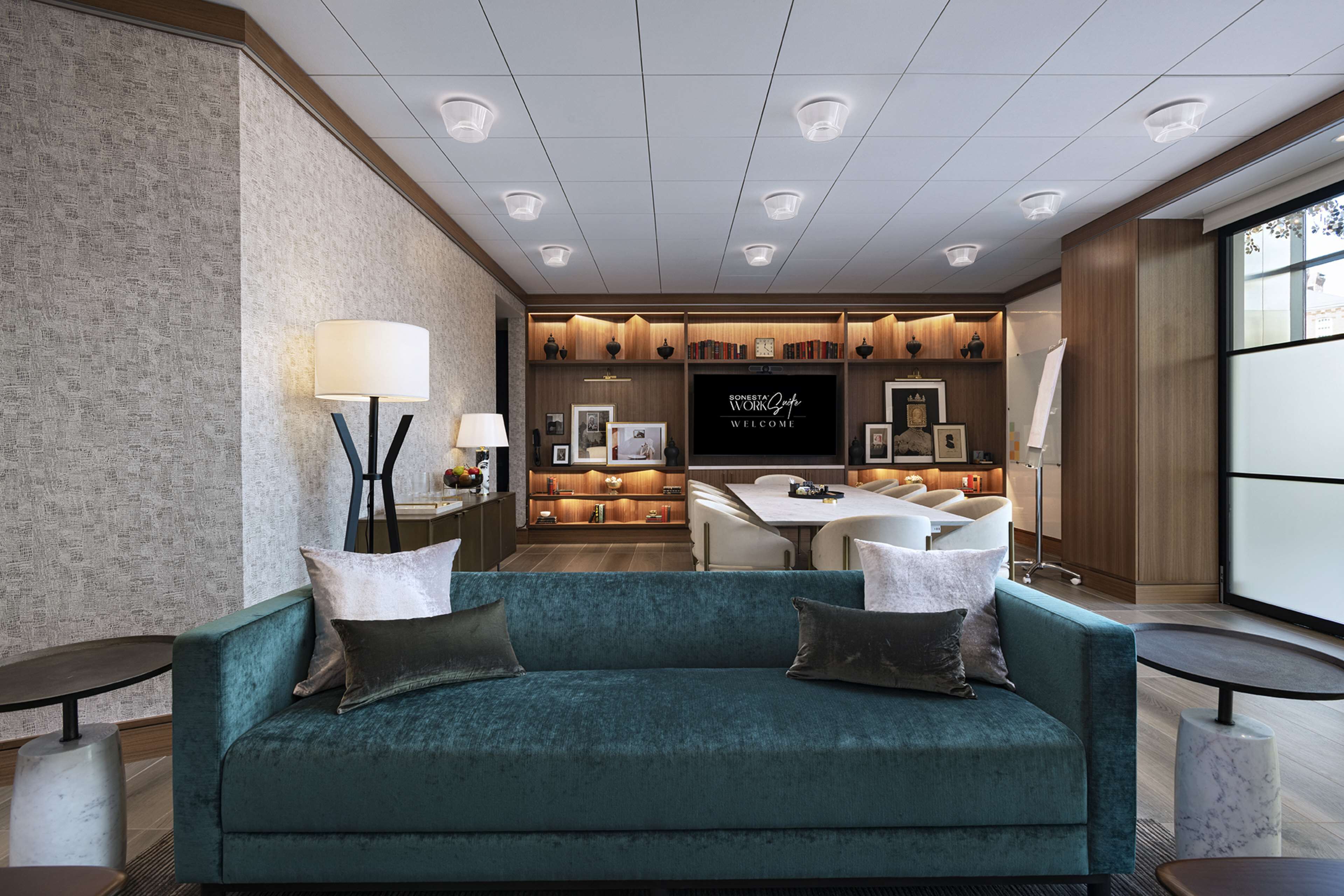 Um hall moderno de hotel com um sofá verde-água, uma área de refeições e uma tela de TV exibindo "WELCOME".