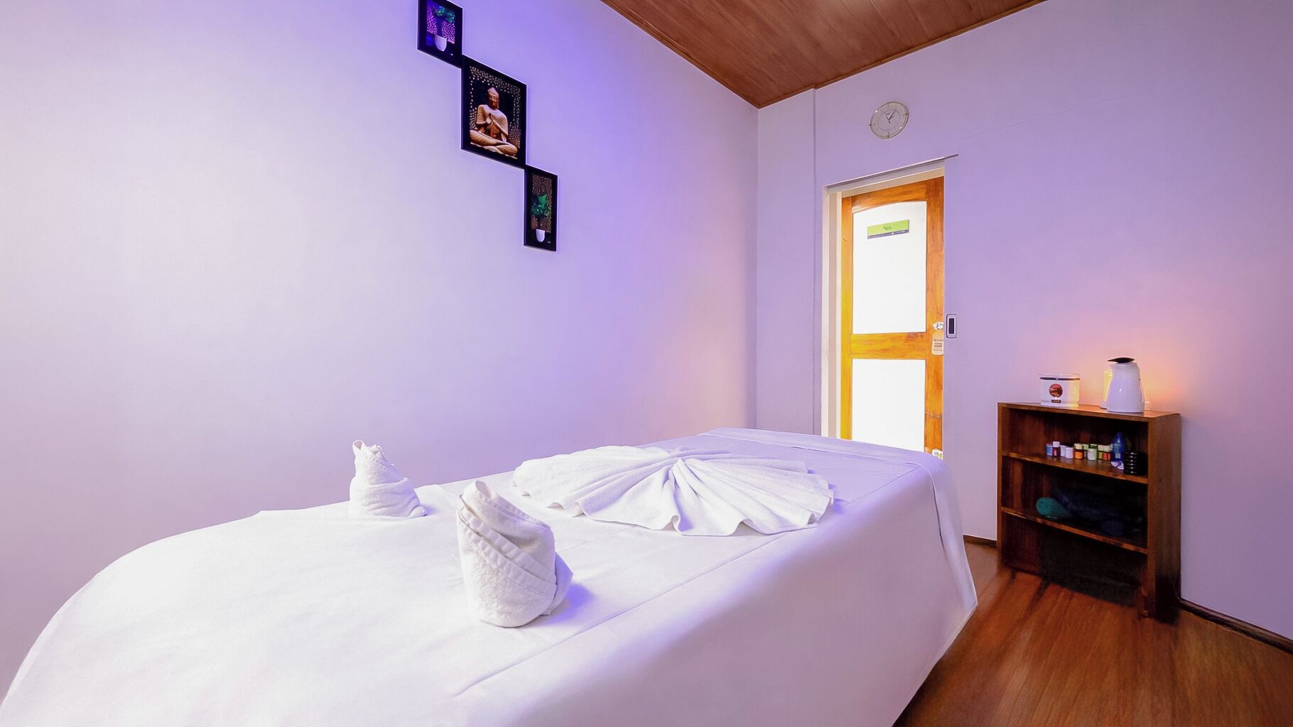 Quarto com uma cama, prateleira de madeira com artigos de higiene e porta que dá para uma área iluminada.