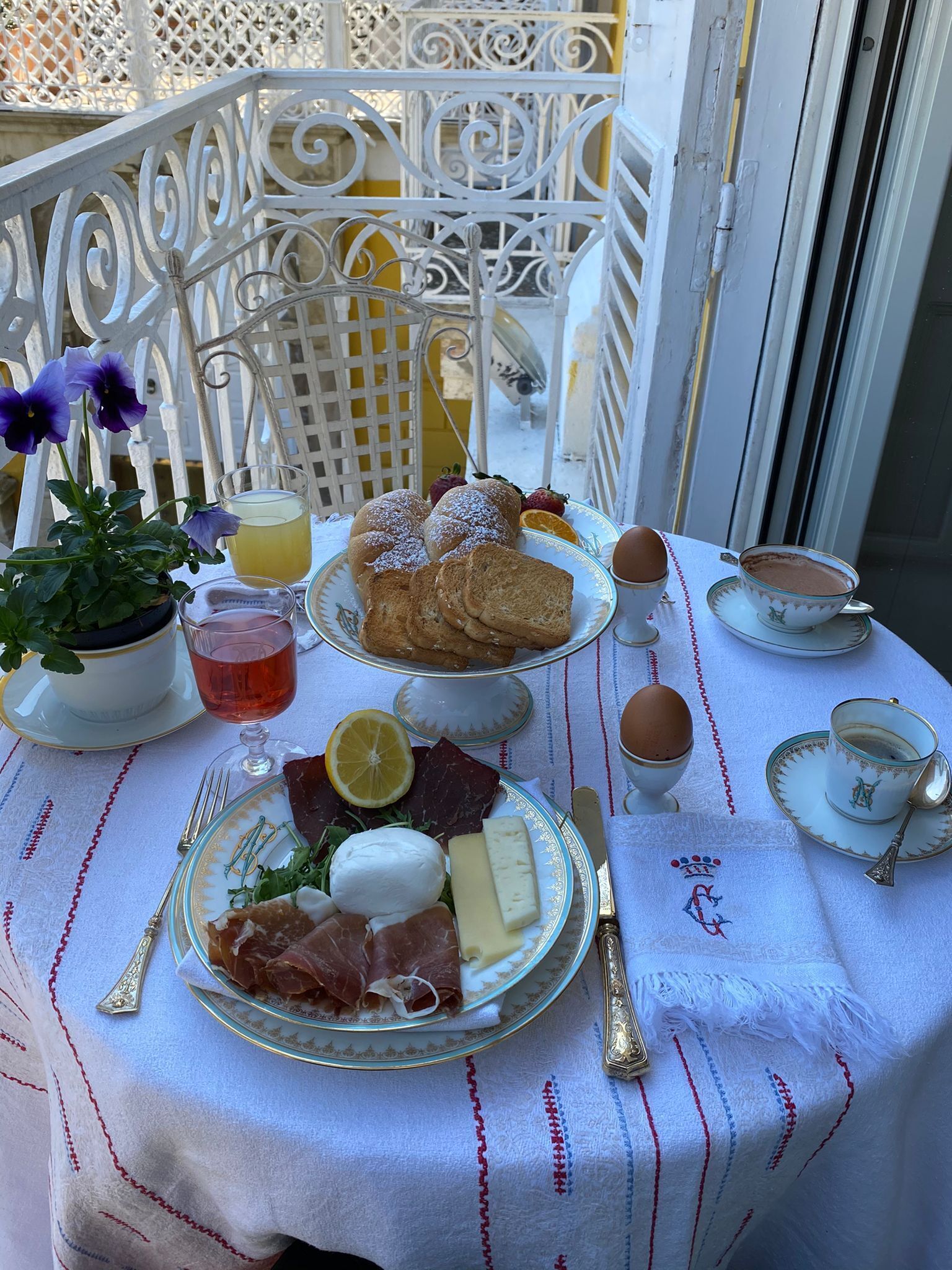 Mesa posta com itens de café da manhã, incluindo doces, frutas, queijo e frios, vista para uma varanda ao fundo.