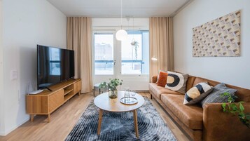 Uma sala de estar moderna com televisão de tela plana, mobiliário de madeira, uma mesa de centro redonda e uma grande janela com cortinas.