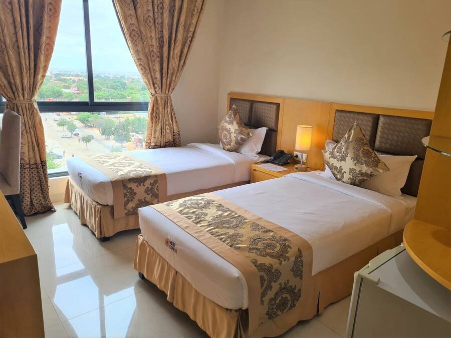 Quarto de hotel com duas camas, uma mesa de cabeceira com abajur e vista da cidade através de grandes janelas.