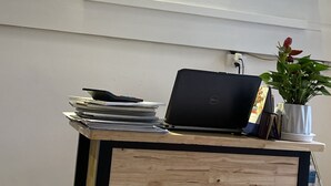 Uma mesa de recepção com um laptop, uma planta e uma pilha de documentos. Há dois relógios na parede acima.
