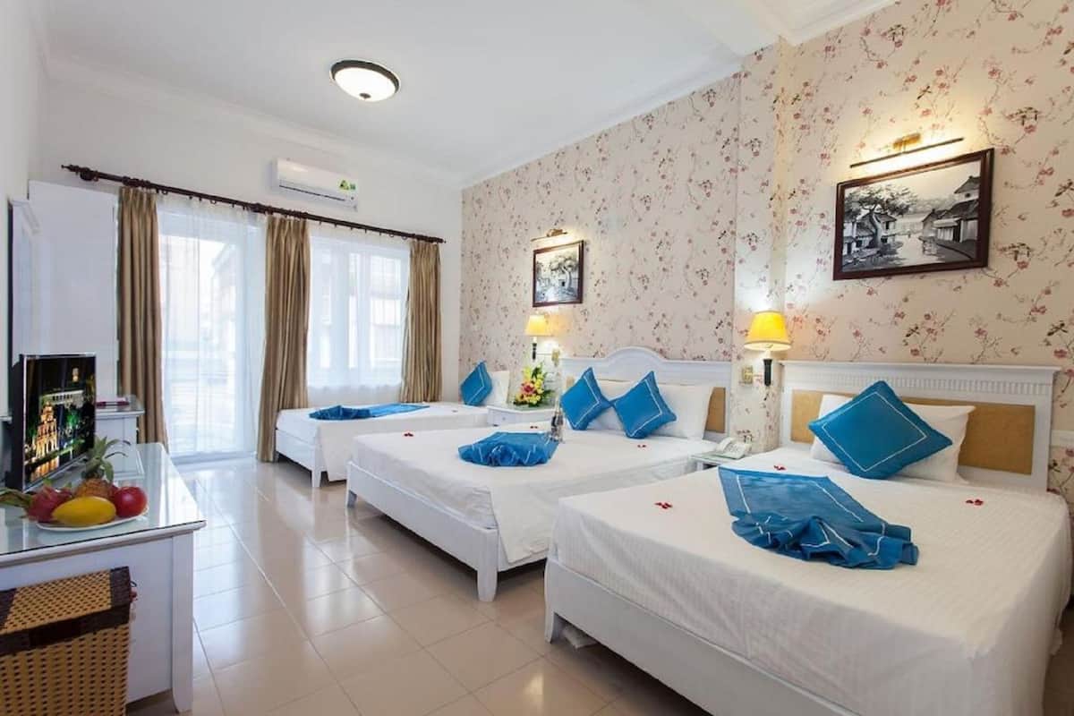 Quarto de hotel com duas camas de solteiro, uma secretária pequena com um computador e uma fruteira, além de uma janela grande com cortinas.