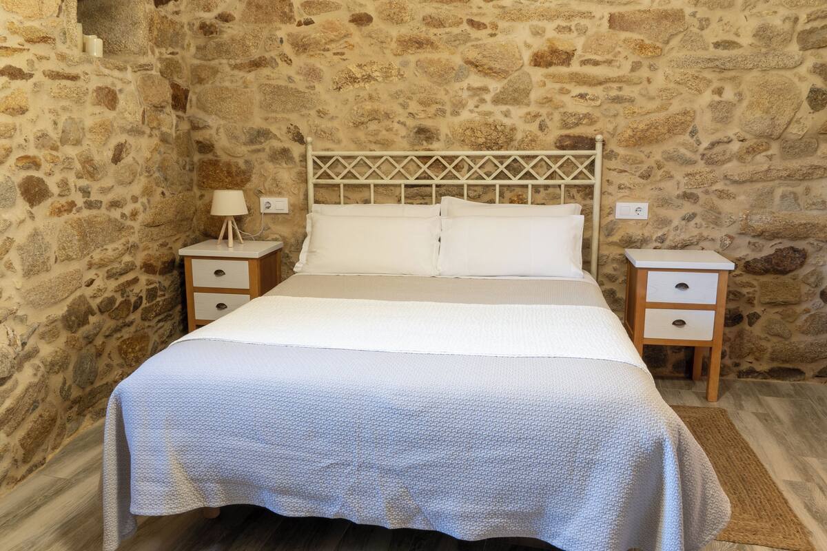 Uma cama com cabeceira branca, duas mesas de cabeceira e um painel de parede de pedra.