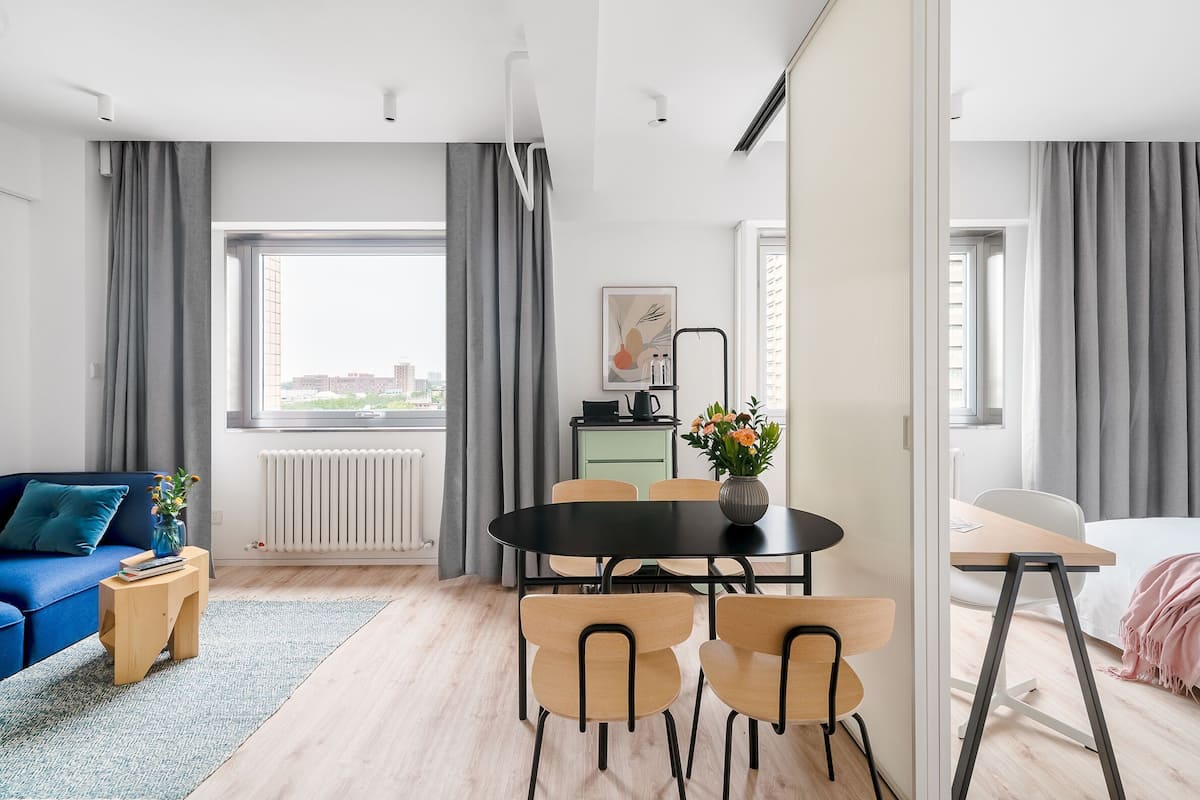 Uma sala de estar moderna com um sofá, uma mesa de jantar com cadeiras, uma mesinha lateral, um radiador e uma janela com cortinas.