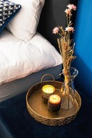 Uma cama com almofadas brancas, um vaso com flores secas e duas velas acesas sobre uma bandeja.