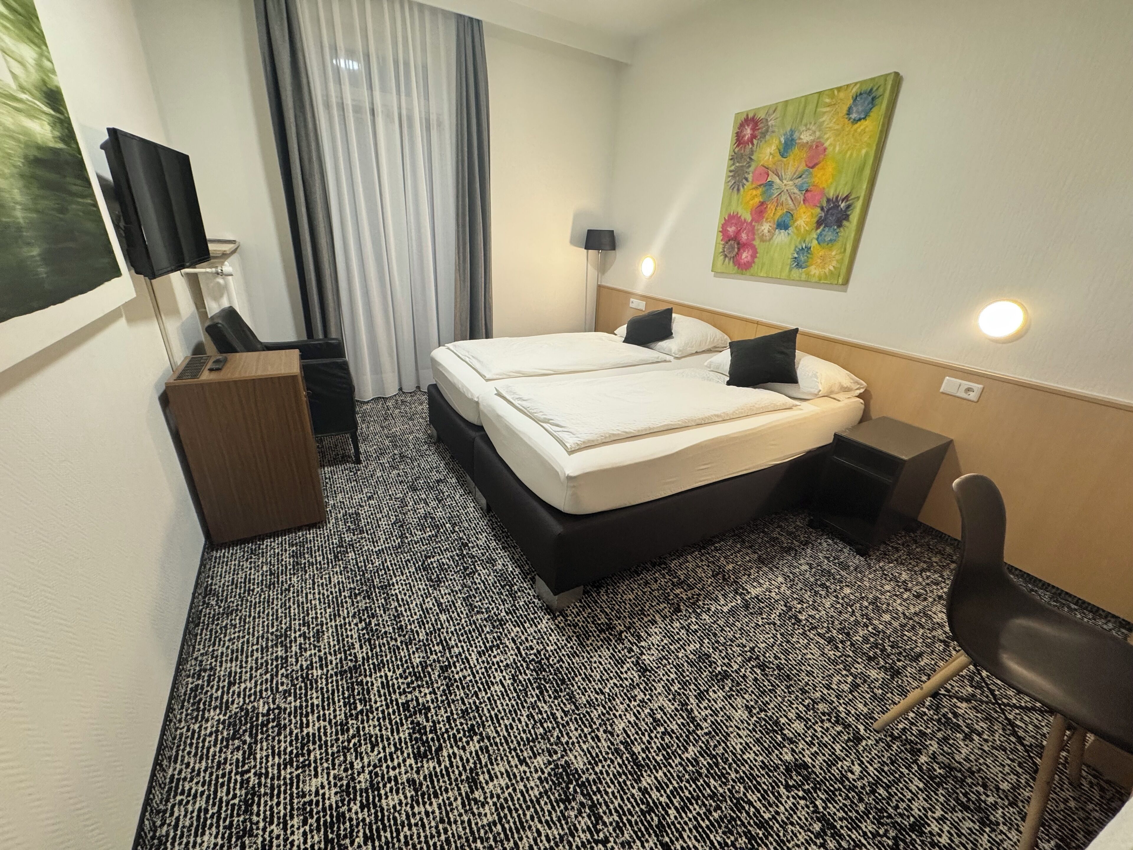 Quarto de hotel com uma cama grande, escrivaninha, cadeira, televisão e um quadro na parede.