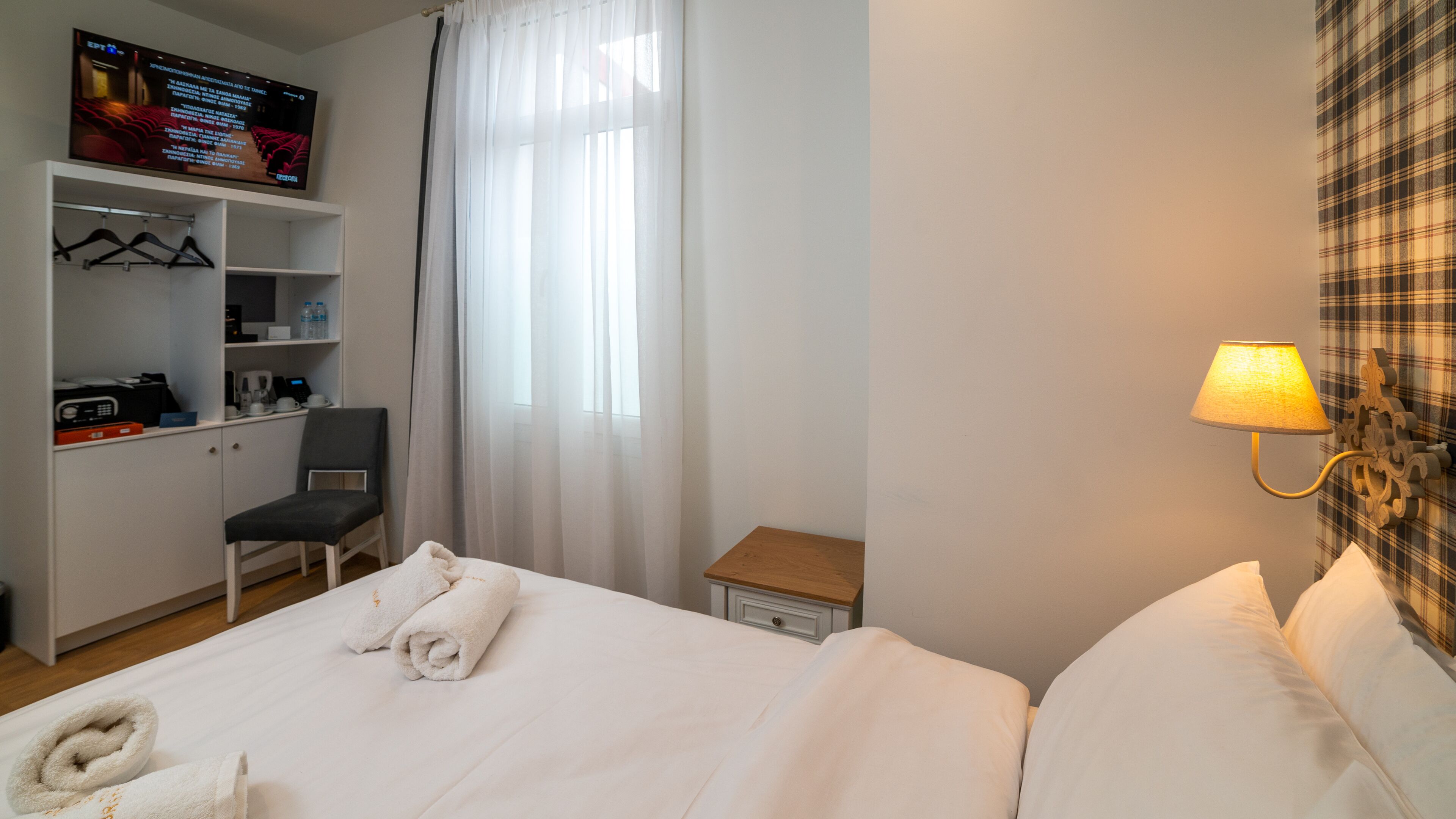 Quarto de hotel com uma cama, uma escrivaninha com candeeiro, uma cadeira, um guarda-roupa e uma televisão na parede.