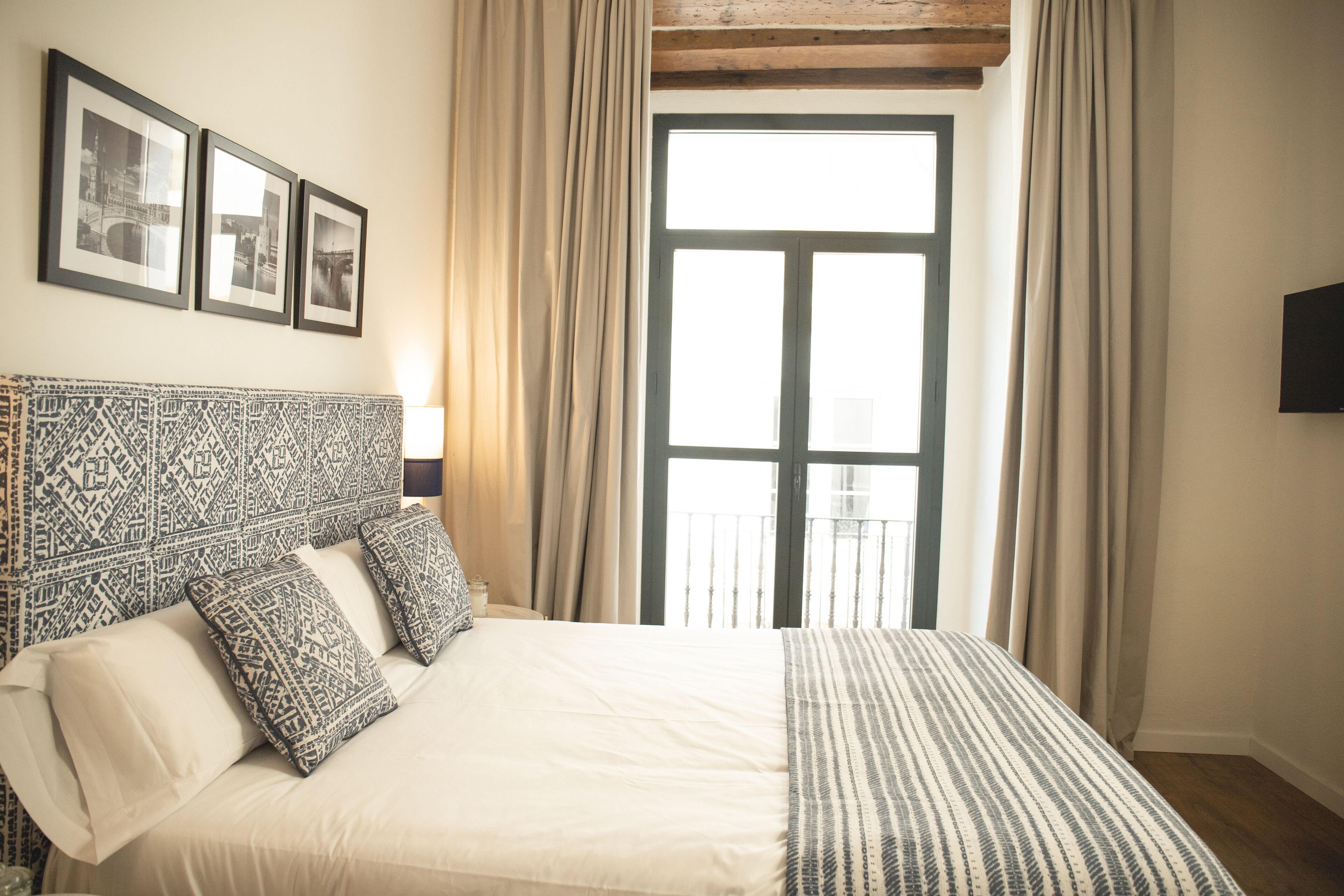 Cama bem arrumada com almofadas estampadas, cabeceira com design geométrico e uma janela grande com cortinas.