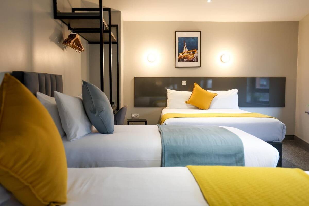 Quarto de hotel com duas camas, cabeceira, mesa de cabeceira, obra de arte na parede e uma escada.