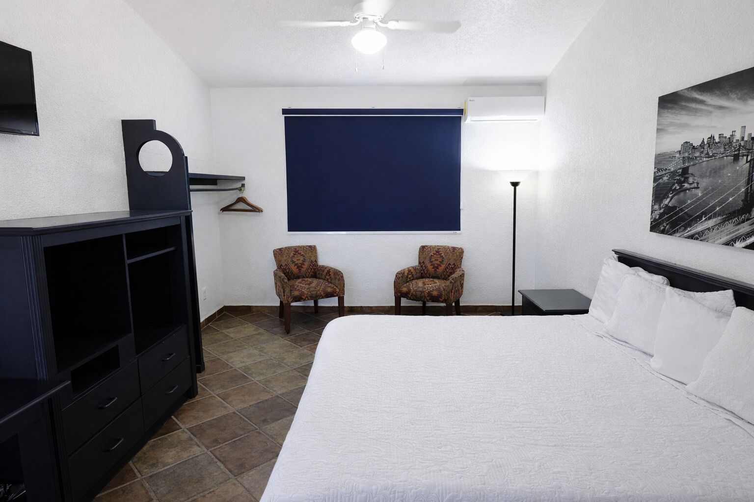 Quarto de hotel com uma cama, duas poltronas, uma televisão, uma escrivaninha e um quadro na parede.