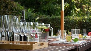 Mesa de madeira posta com copos de vinho transparentes e uma garrafa, cercada por vegetação.