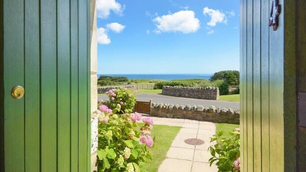 Vista de uma porta verde para um jardim com flores cor-de-rosa e um muro de pedra.