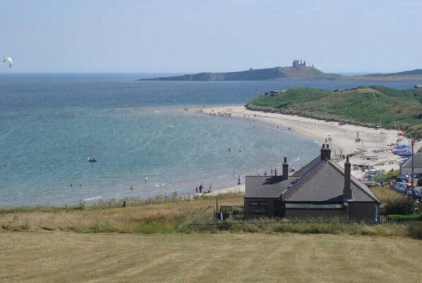 Vista costeira com praia, um castelo numa colina e um edifício em primeiro plano.
