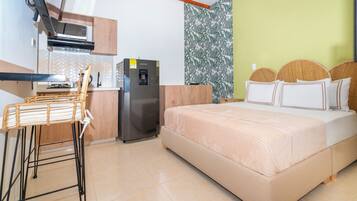 Quarto de hotel compacto com cozinha americana, uma cama com cobertor bege e parede verde decorativa.
