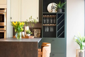 Uma cozinha moderna com bancada escura, armários brancos e uma estante verde com copos de vinho e plantas.