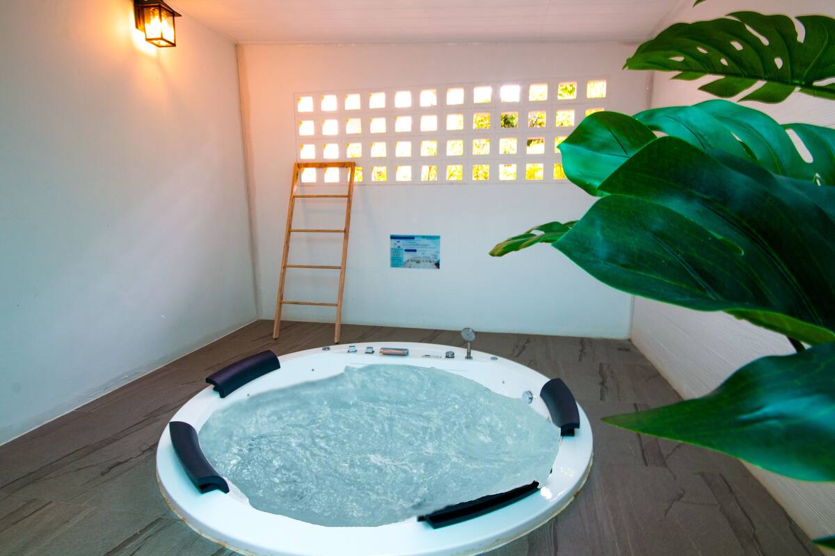 Uma banheira de hidromassagem em um quarto com piso de madeira, uma escada, parede com uma grade de janelas pequenas e uma grande planta verde.