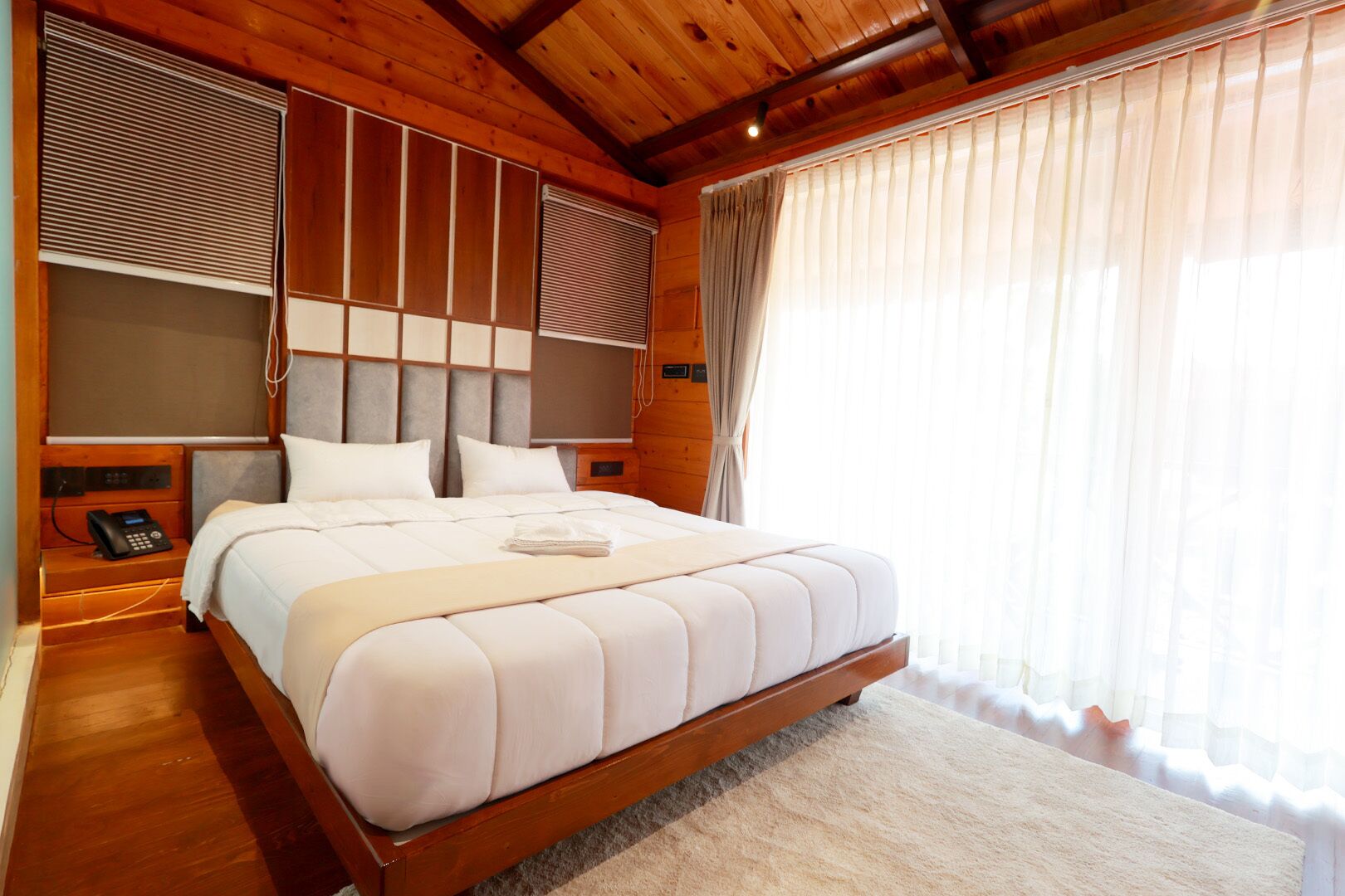 Uma cama grande com cabeceira de madeira, telefone ao lado da cama e janela com cortinas translúcidas.