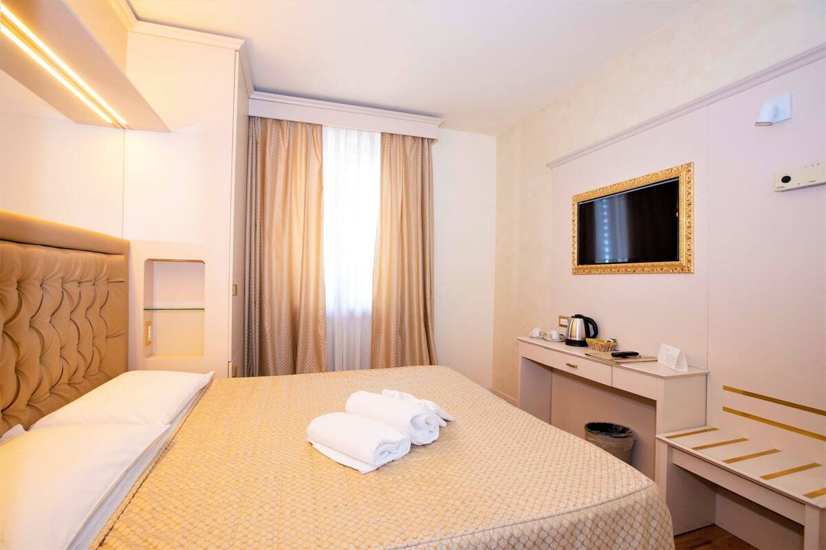 Quarto de hotel com cama, mesa de cabeceira, escrivaninha, espelho e janela com cortinas.