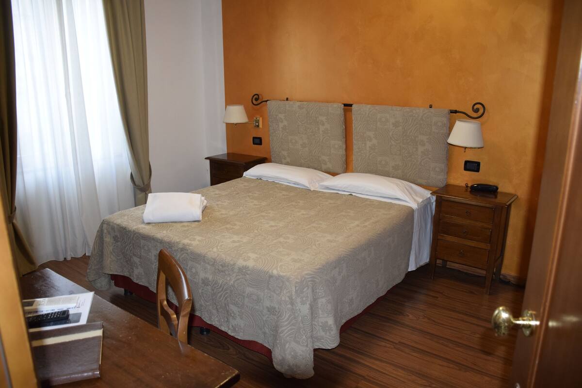 Uma cama bem arrumada com colcha estampada, duas mesas de cabeceira com luminárias, uma cadeira de madeira e uma escrivaninha de madeira com um livro e um controle remoto.
