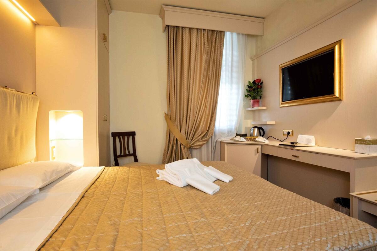 Quarto de hotel com cama, escrivaninha, cadeira, televisão e janela com cortinas.