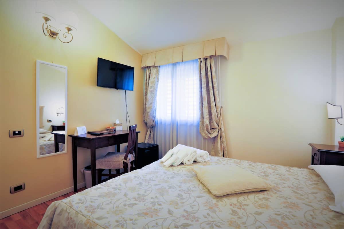 Quarto de hotel com uma cama, uma mesa de trabalho com cadeira, televisão e janela com cortinas.