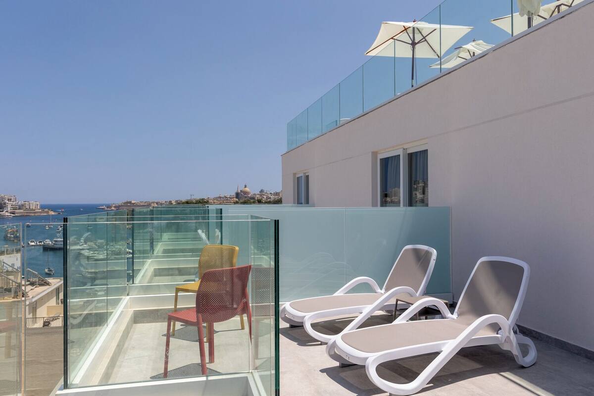 Um terraço no telhado com guarda-corpos de vidro, uma cadeira amarela e duas cadeiras de descanso brancas.
