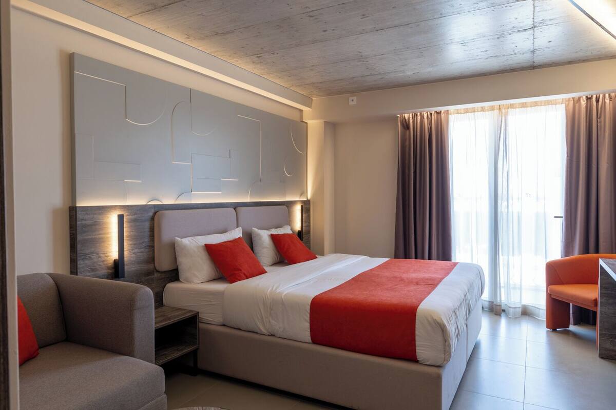 Quarto de hotel moderno com uma cama grande, um sofá, uma escrivaninha e vista para o exterior pela janela.