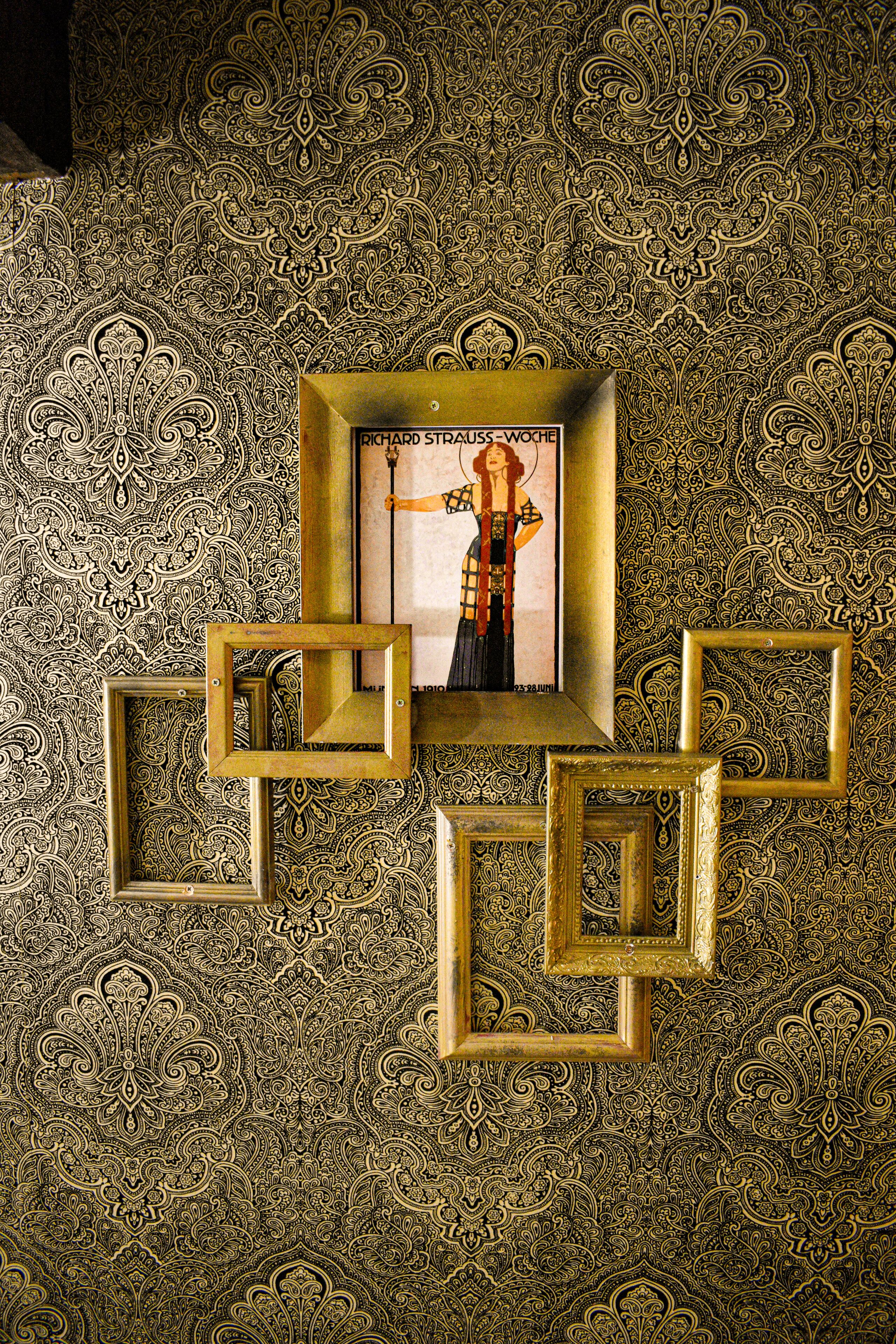 Paredes com papel de parede ornamentado em padrão dourado e um quadro com uma mulher num vestido.