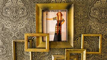 Paredes com papel de parede ornamentado em padrão dourado e um quadro com uma mulher num vestido.
