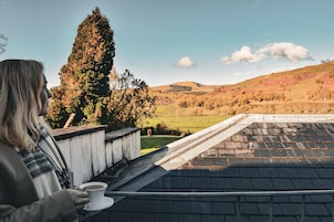 Uma pessoa segurando uma xícara em um terraço com vista para uma paisagem montanhosa.