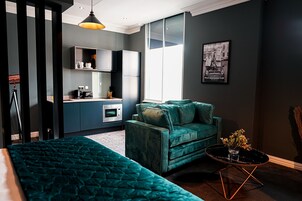 Um quarto moderno com uma cama grande, um sofá verde-água, uma pequena mesa com um vaso e uma cozinha compacta ao fundo.