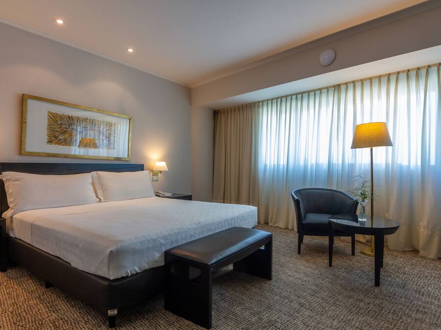 Quarto de hotel com uma cama grande, uma mesa de cabeceira com abajur, uma cadeira e uma janela com cortinas.